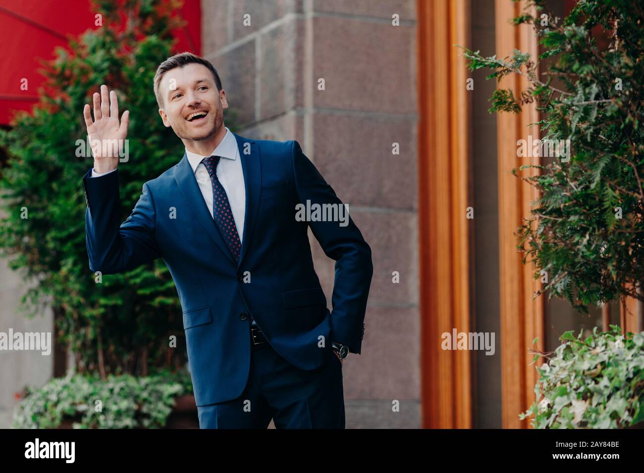 Shot of cheerful male banker in luxury black suit, waves with hand as ...