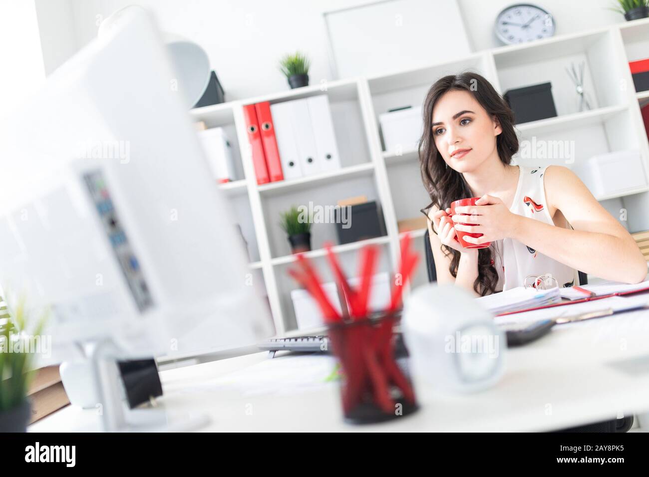 Beautiful young girl sits at office desk, looks at computer screen and ...