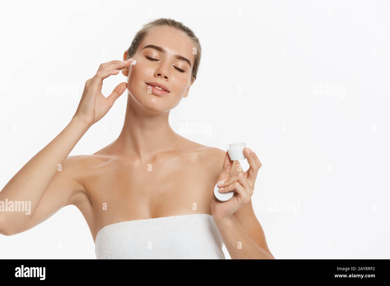 Beautiful young woman applying cosmetic cream treatment on her face isolated on white background ...