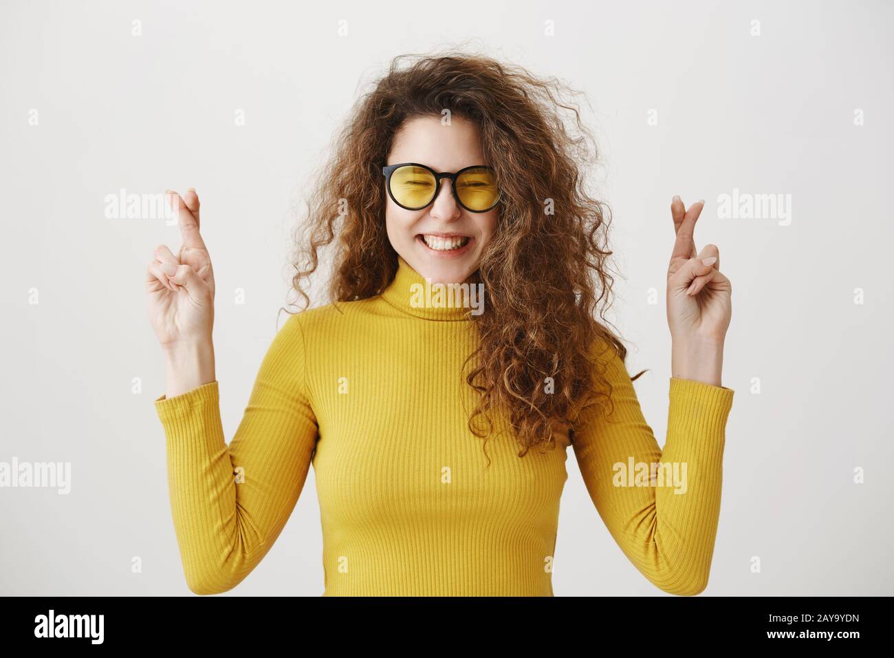 Excited woman in yellow sweater keeping fingers crossed, mouth wide ...