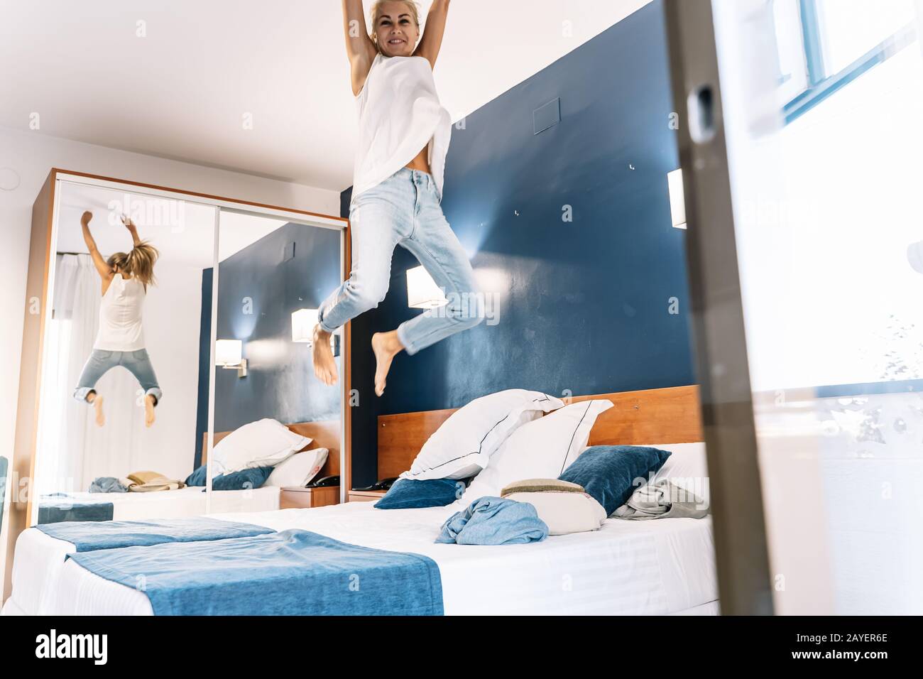Blonde girl jumping on a hotel bed with the blue sheets and a mirror ...