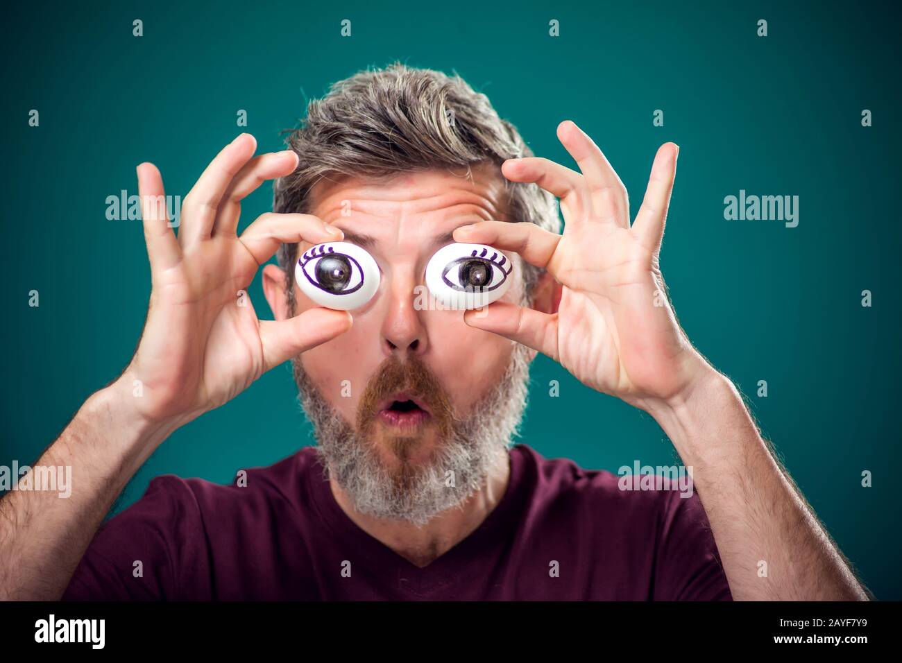 A portrait of funny bearded man in red t-shirt holding eggs behind eyes ...