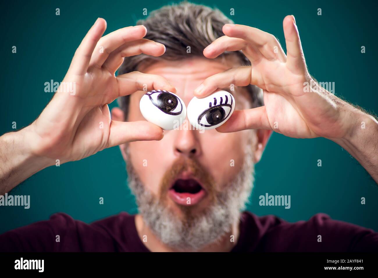 A portrait of funny bearded man in red t-shirt holding eggs behind eyes ...