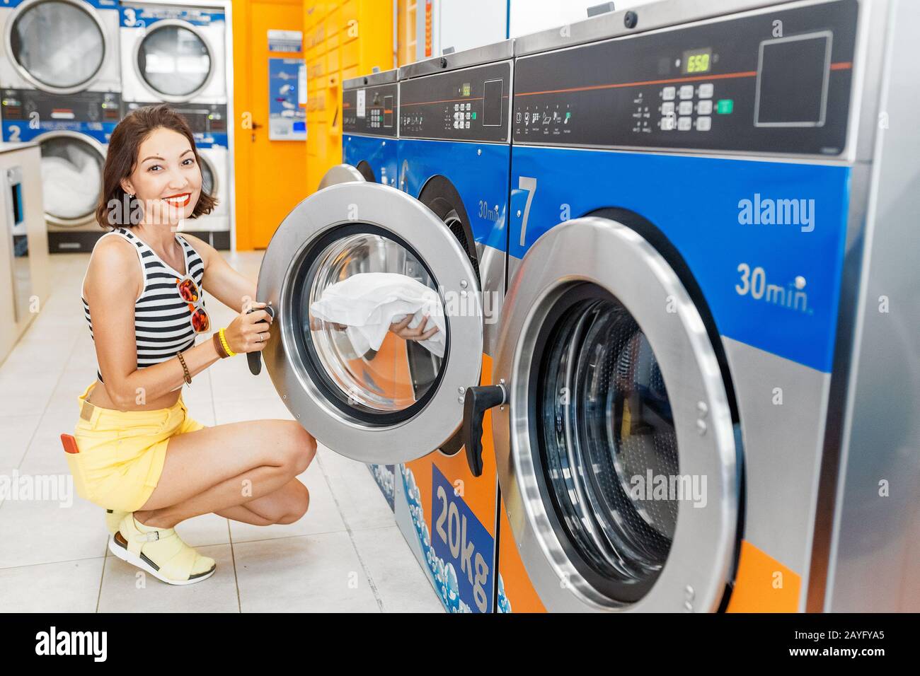 Young woman loading dirty clothes in washing machine in laundromat ...