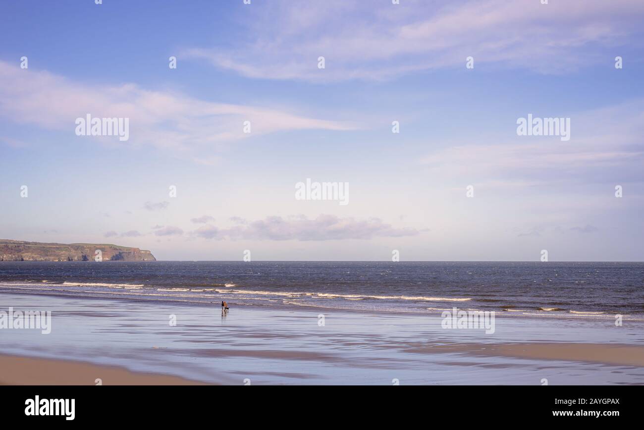 Whitby beach. Waves roll onto a wet beach as a couple walk their dog. A ...