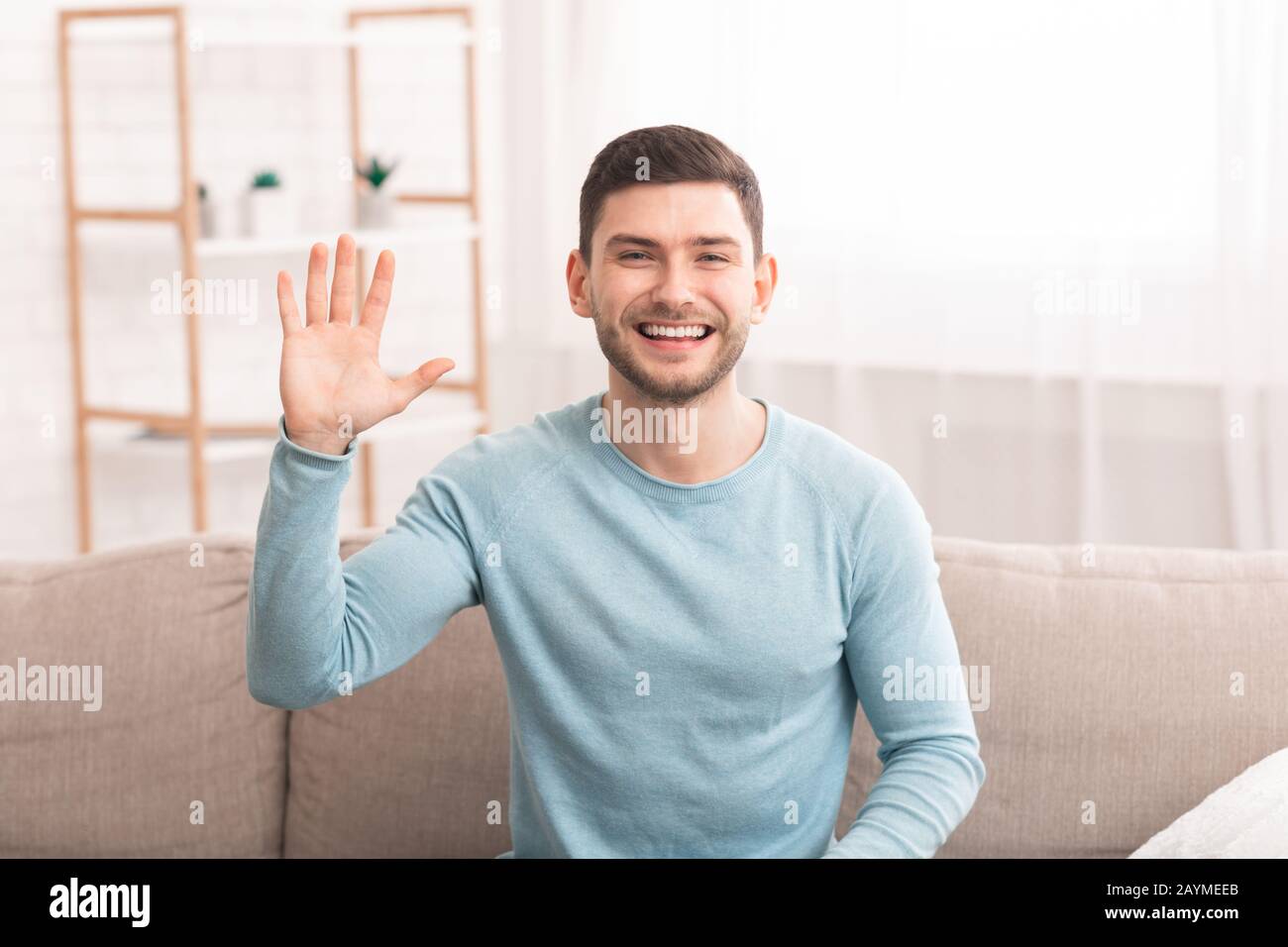 Millennial Guy Waving Hello Smiling To Camera Sitting On Sofa Stock ...