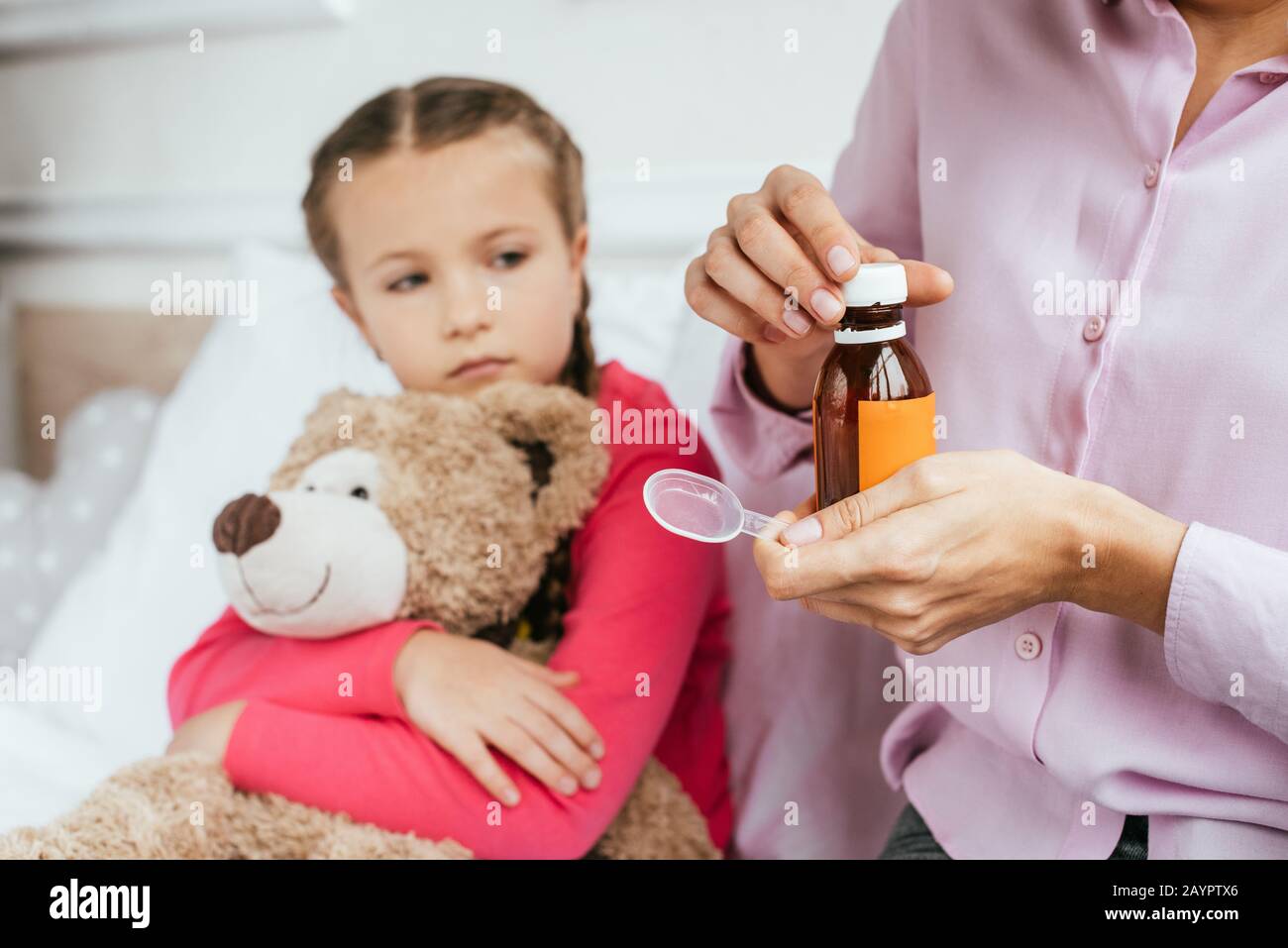 cropped view of mother giving syrup to ill daughter with teddy bear ...