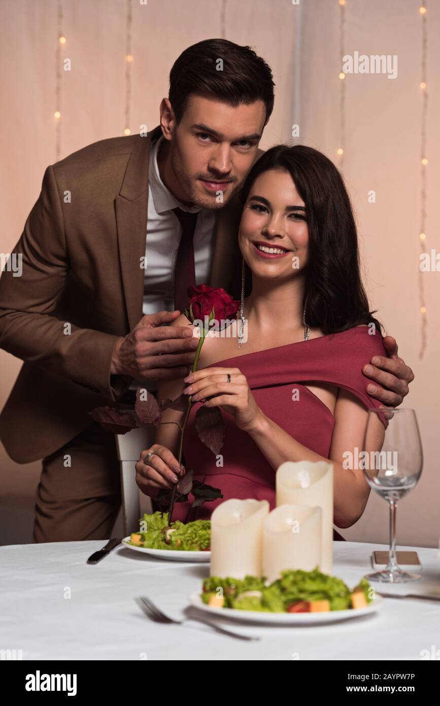 handsome man hugging happy girlfriend holding red rose while looking at ...