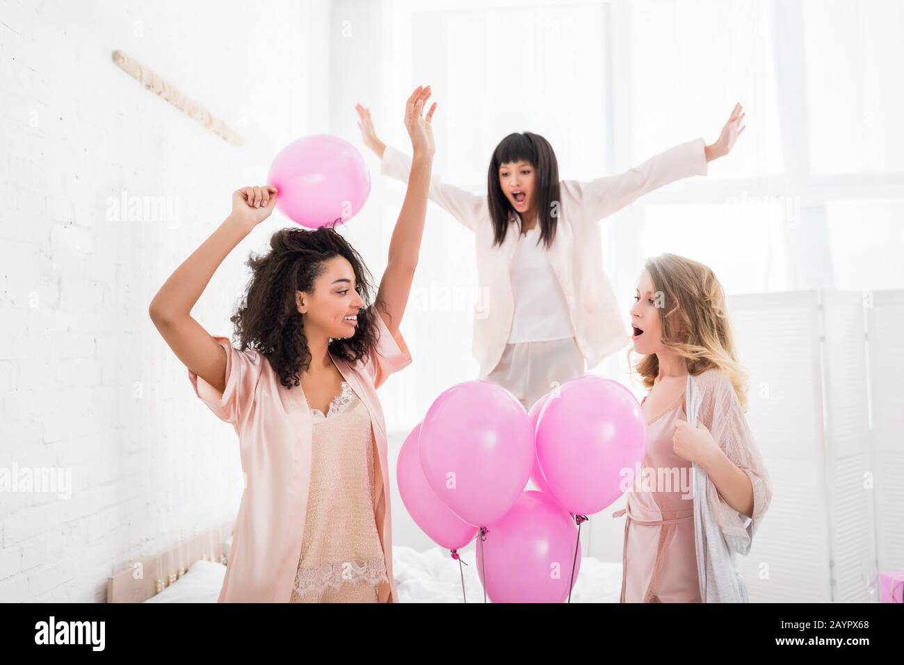 attractive happy multicultural girlfriends dancing and jumping on bed ...