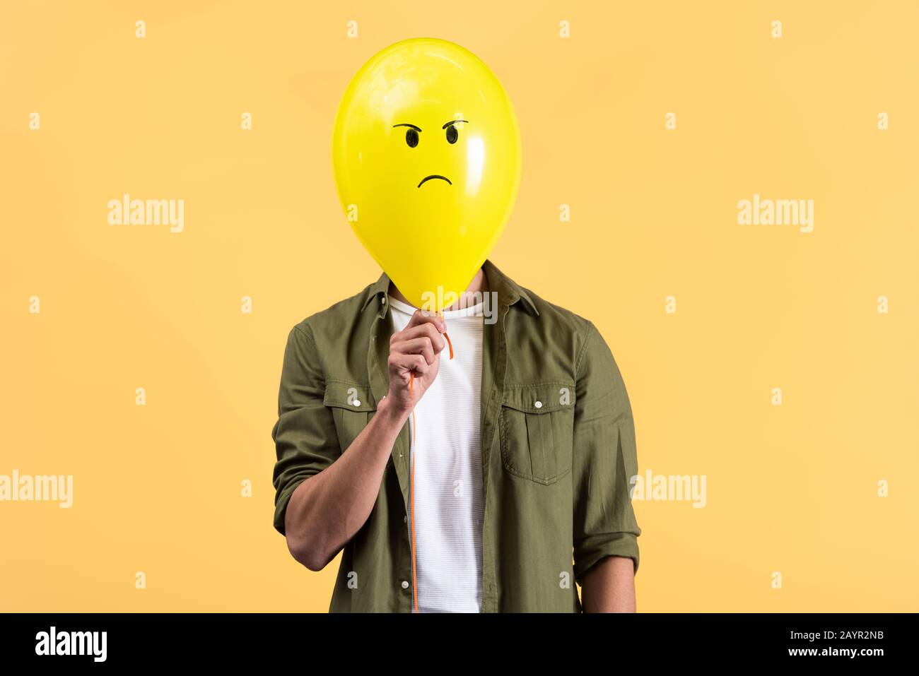 young man holding angry balloon in front of face, isolated on yellow ...