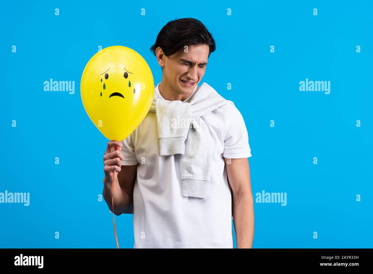 sad young man holding yellow crying balloon, isolated on blue Stock ...
