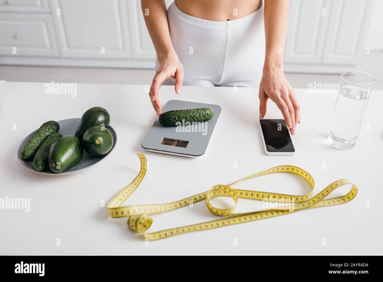 Cropped view of slim woman putting cucumber on scales near smartphone ...
