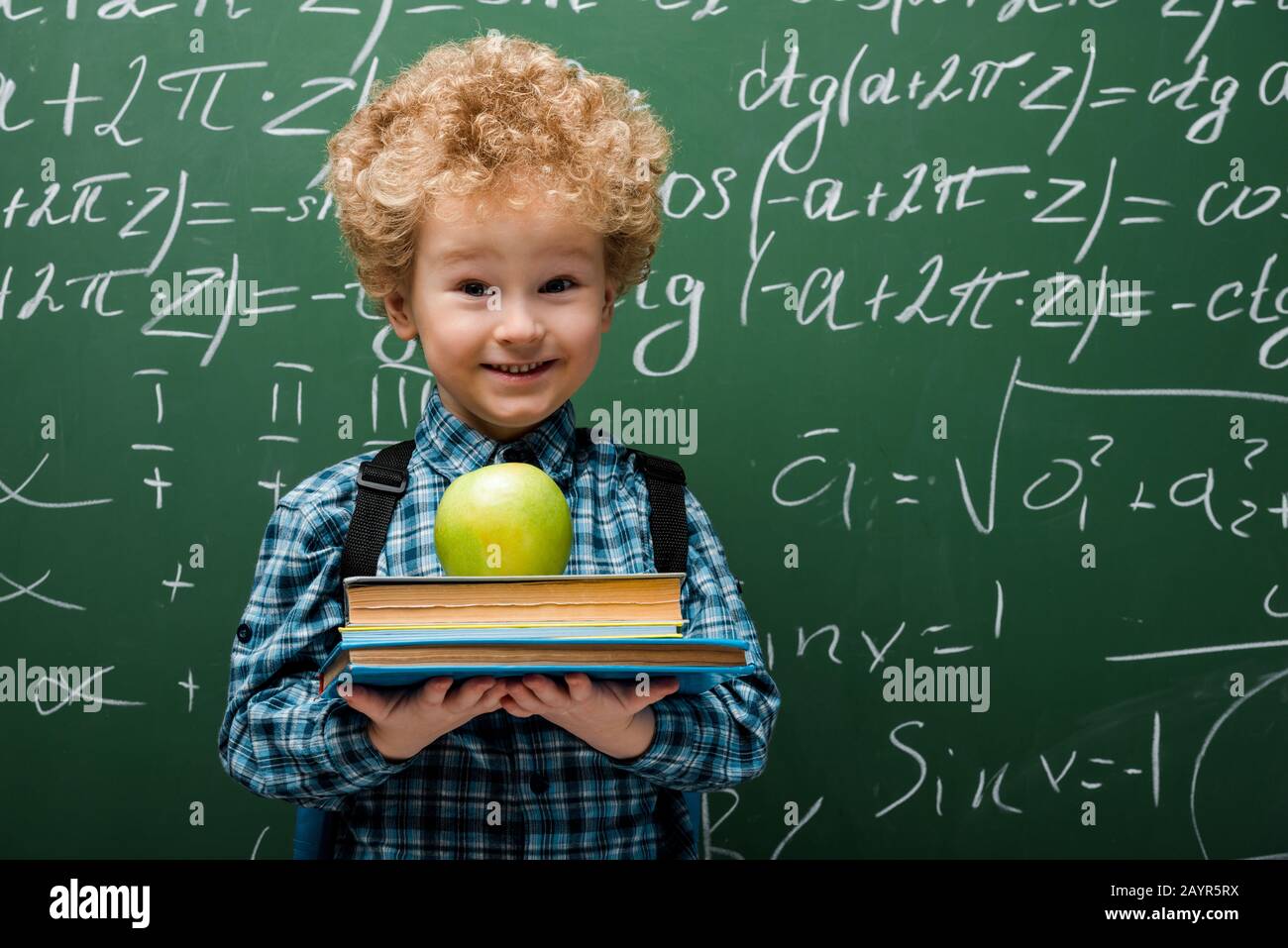 curly kid holding books and apple near chalkboard with mathematical ...
