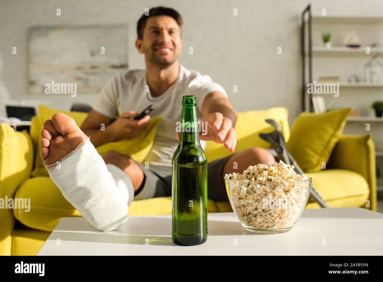 Selective focus of smiling man with broken leg taking beer near popcorn ...