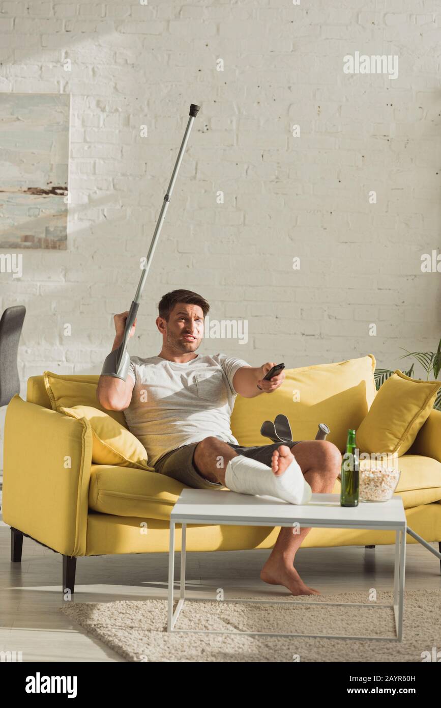 Angry man with broken leg watching tv near beer bottle and popcorn on ...