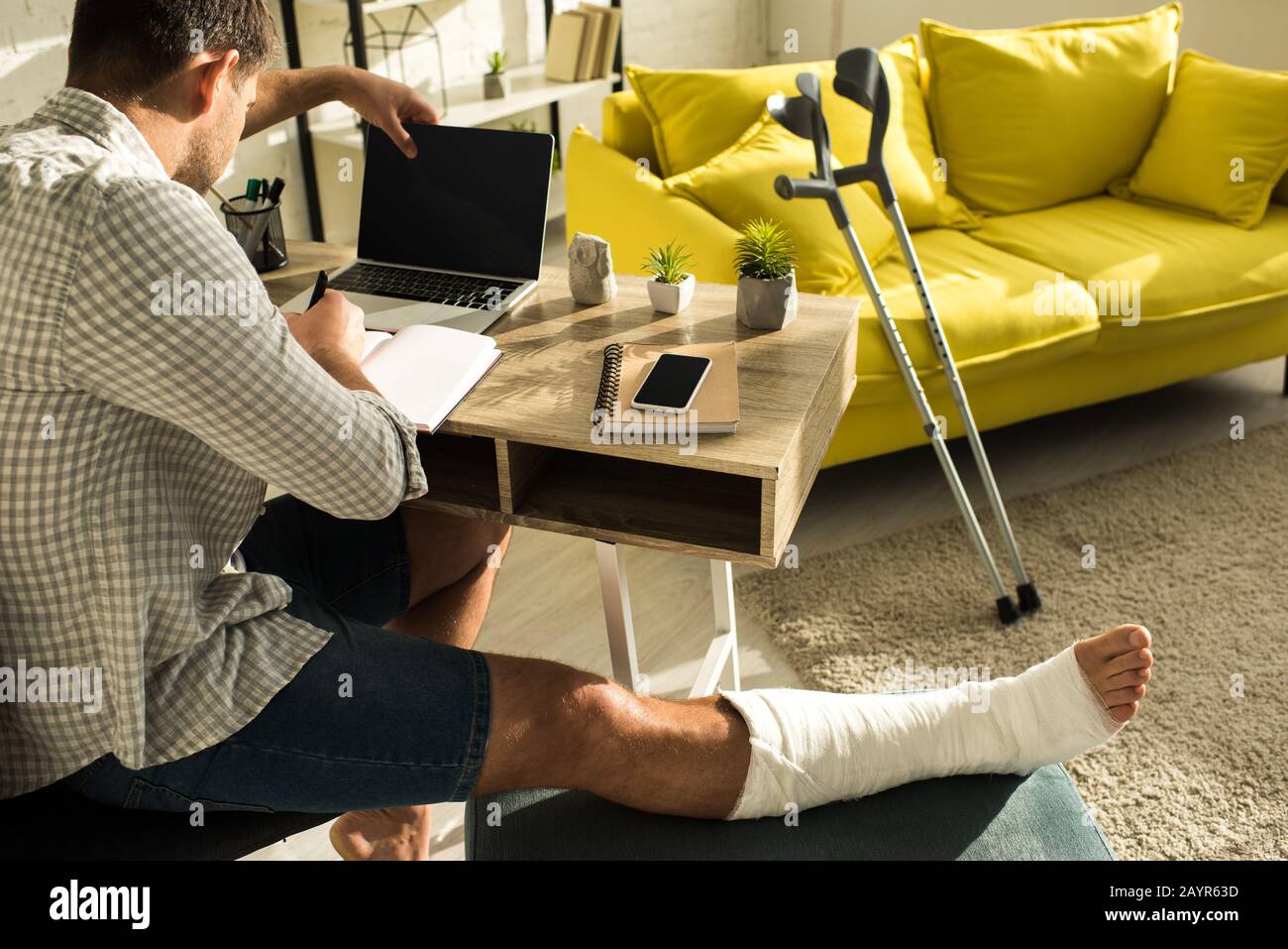 Back view of freelancer with broken leg working with laptop and ...