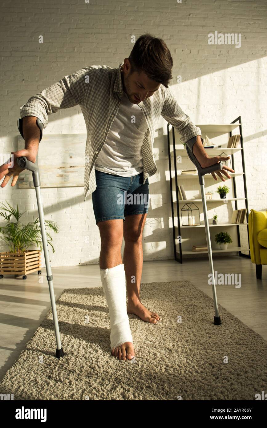 Handsome man with broken leg holding crutches while standing in living ...