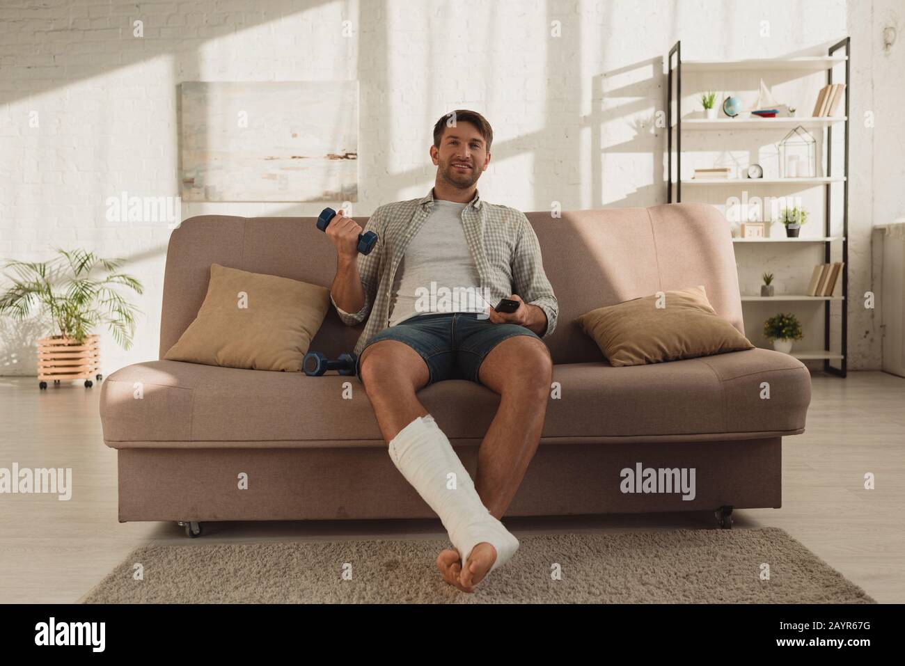 Smiling man with broken leg watching tv and holding dumbbell on sofa at ...