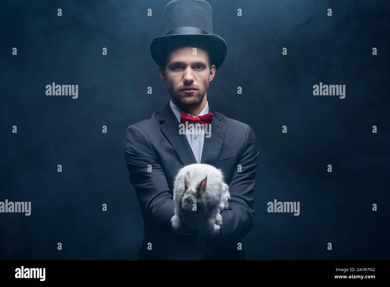 confident magician in suit and hat holding white rabbit, dark room with ...
