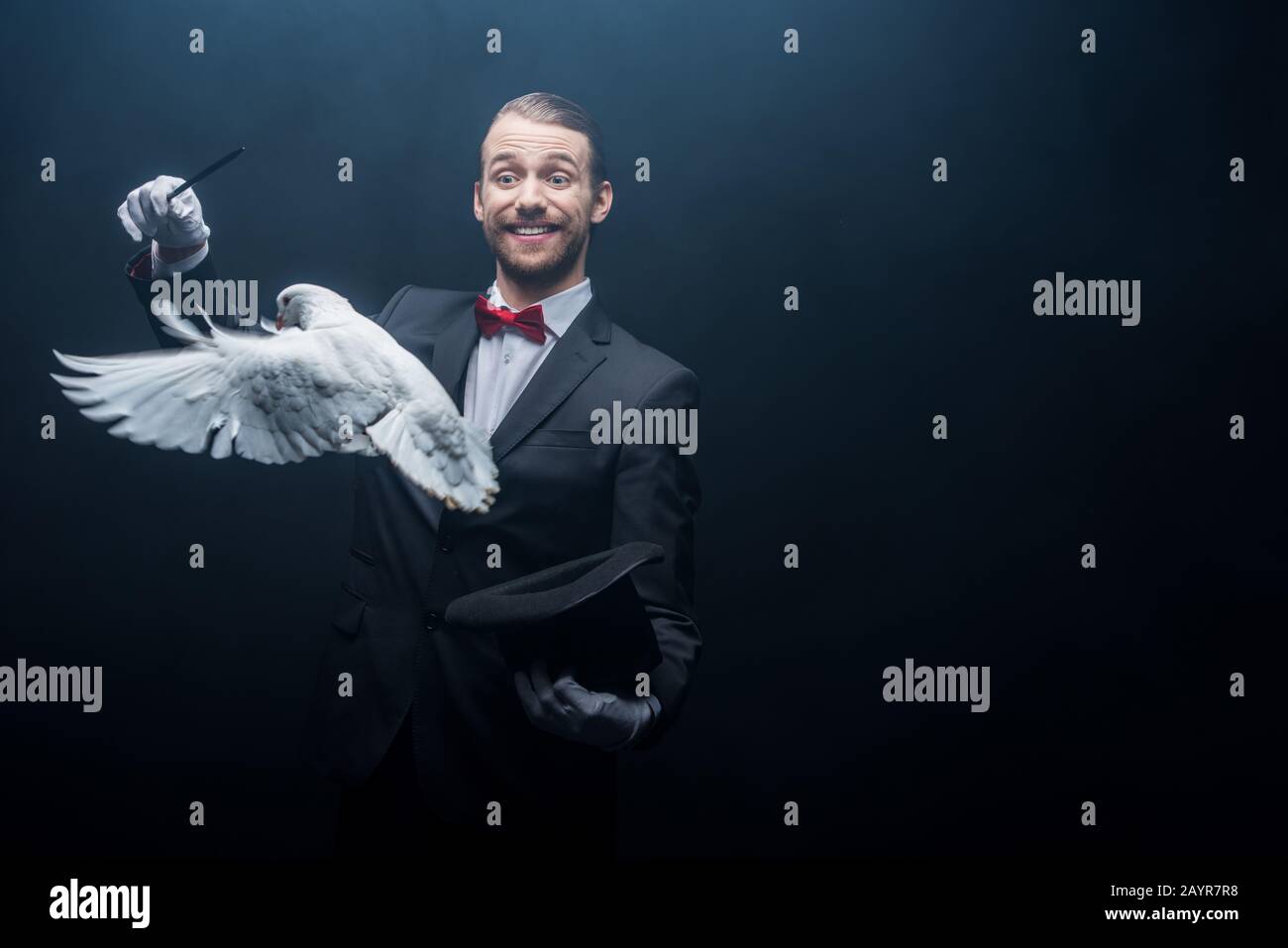 smiling magician showing trick with dove, wand and hat in dark room ...