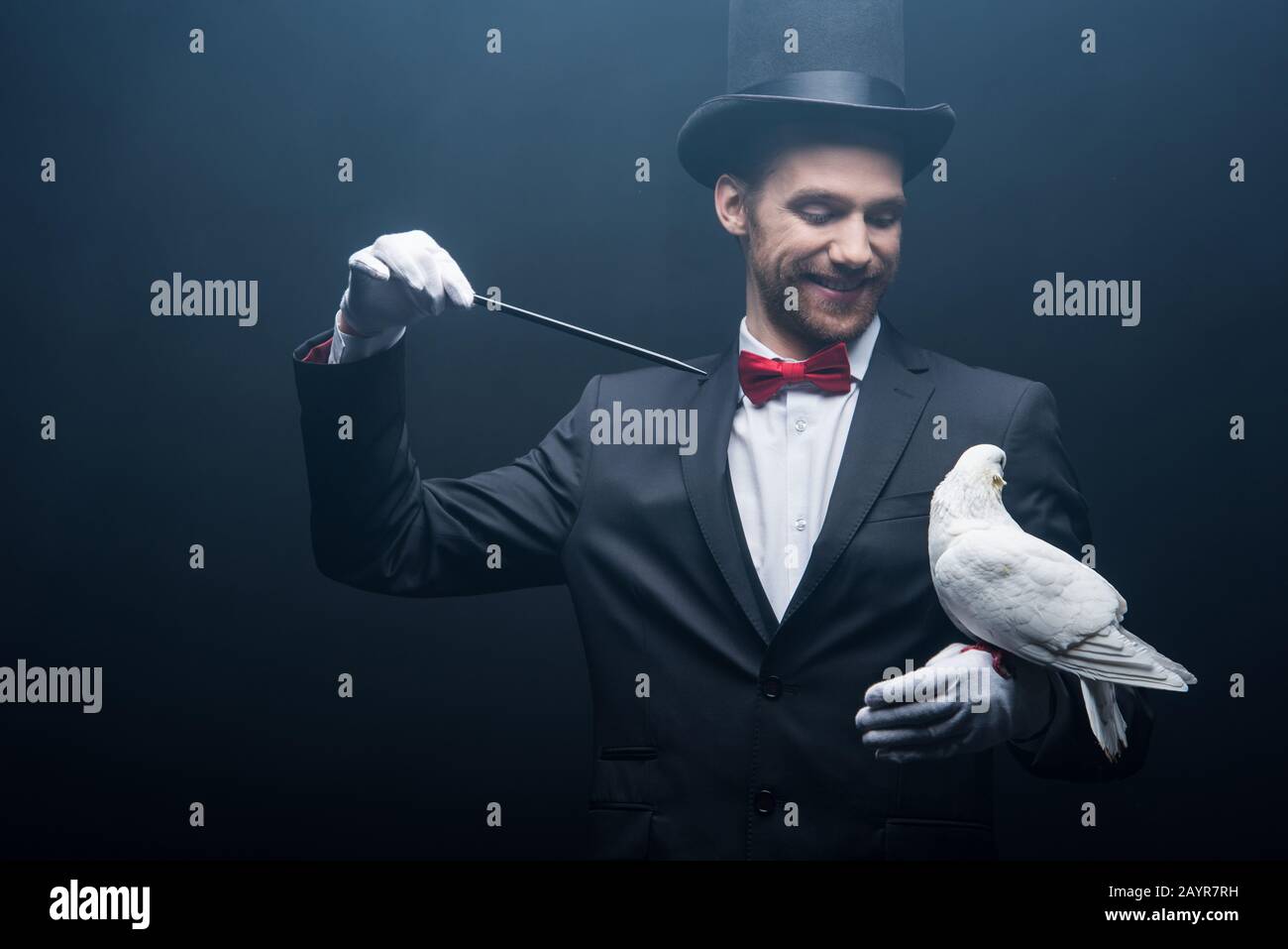 happy magician in hat making abracadabra with dove and wand in dark ...