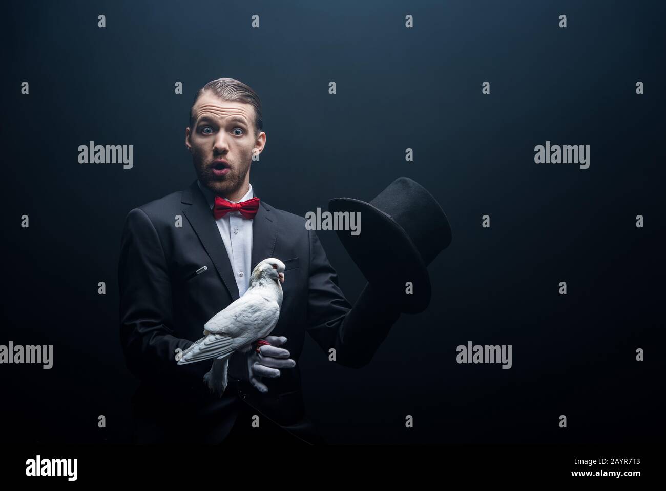 shocked magician showing trick with dove and hat in dark room with ...