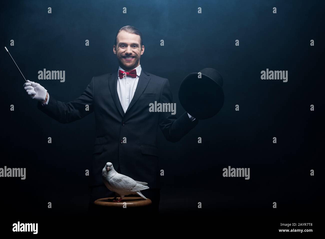happy magician showing trick with dove, wand and hat in dark room with ...