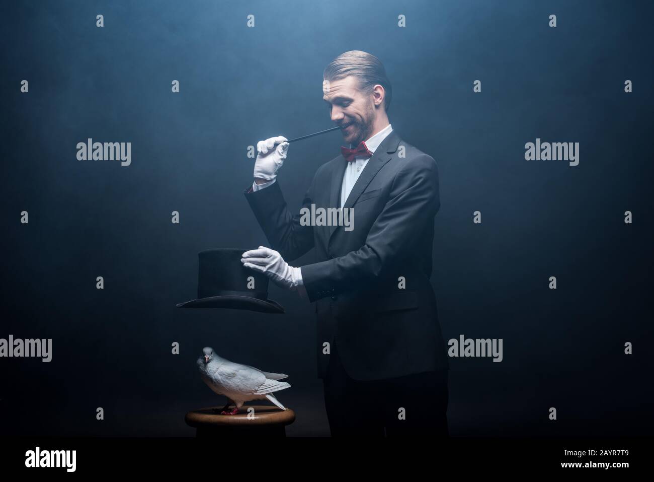 joyful magician showing trick with dove, wand and hat in dark room with ...
