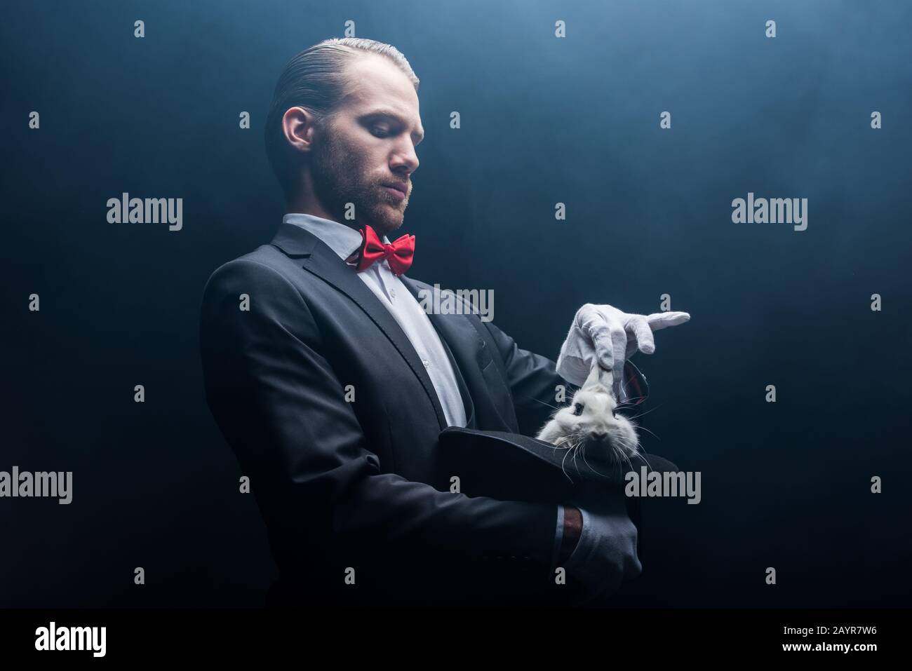 professional magician taking white rabbit from hat, dark room with ...