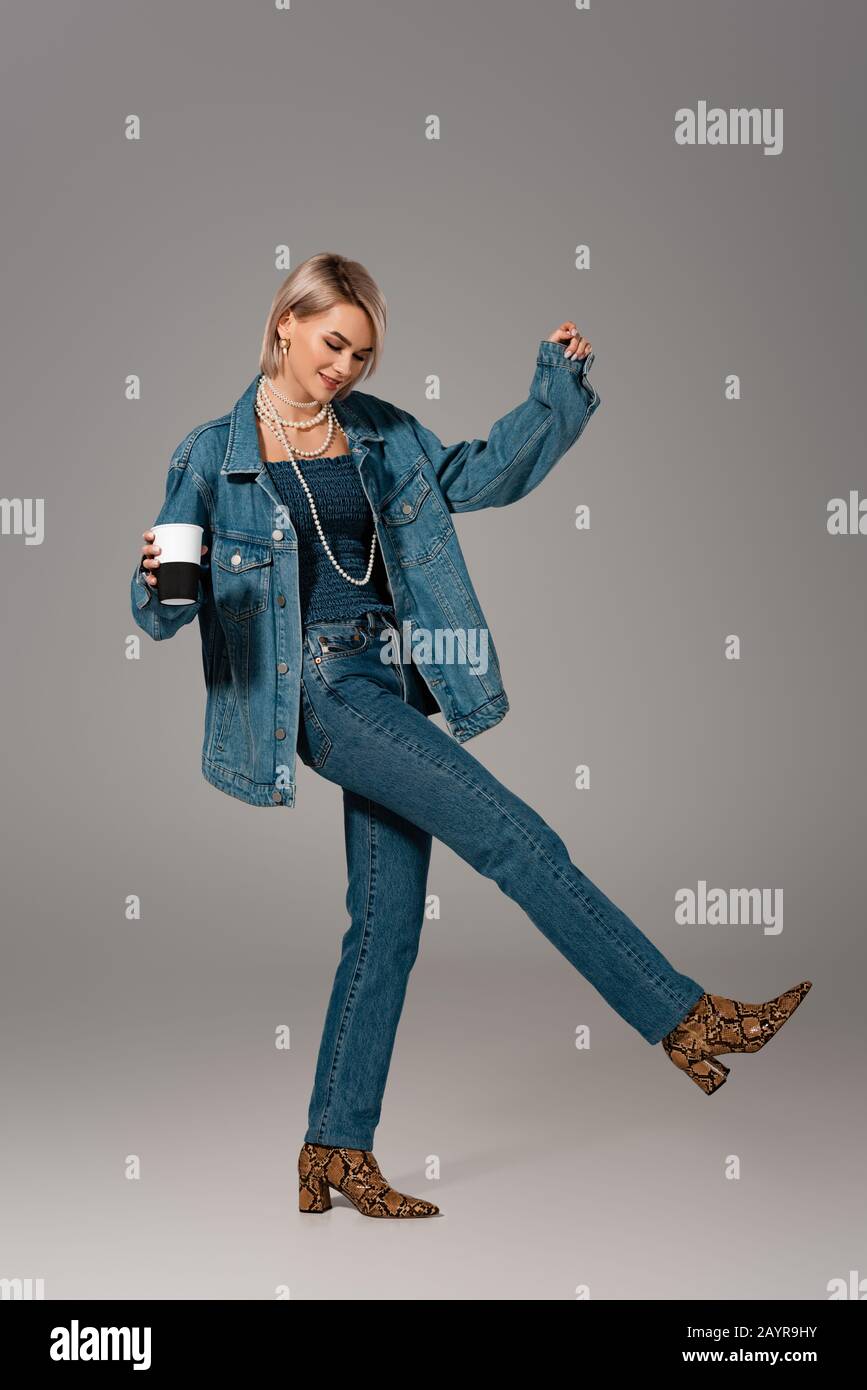 smiling woman in denim jacket and jeans holding insulated mug on grey ...