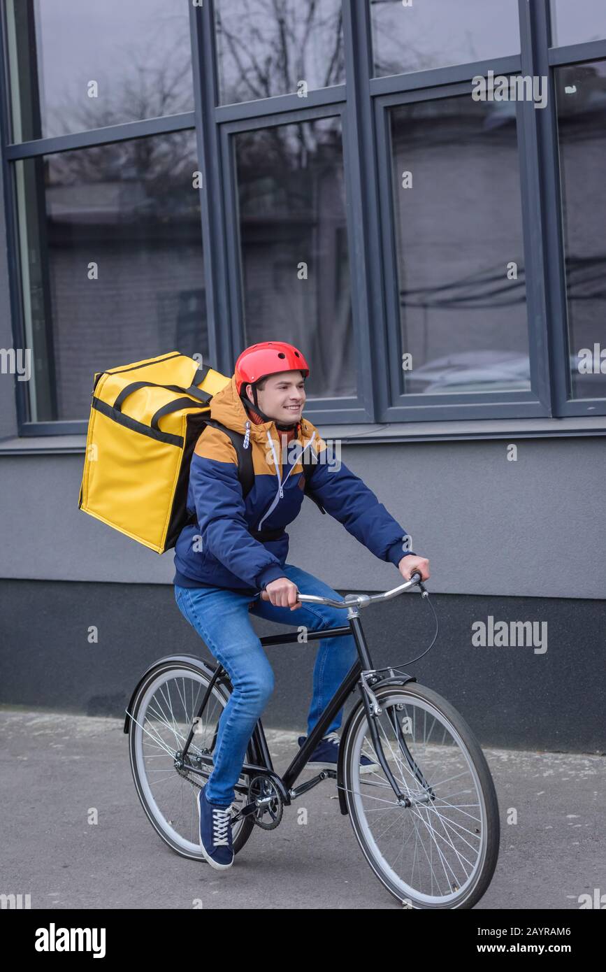 Smiling delivery man with thermo backpack riding bicycle near building ...