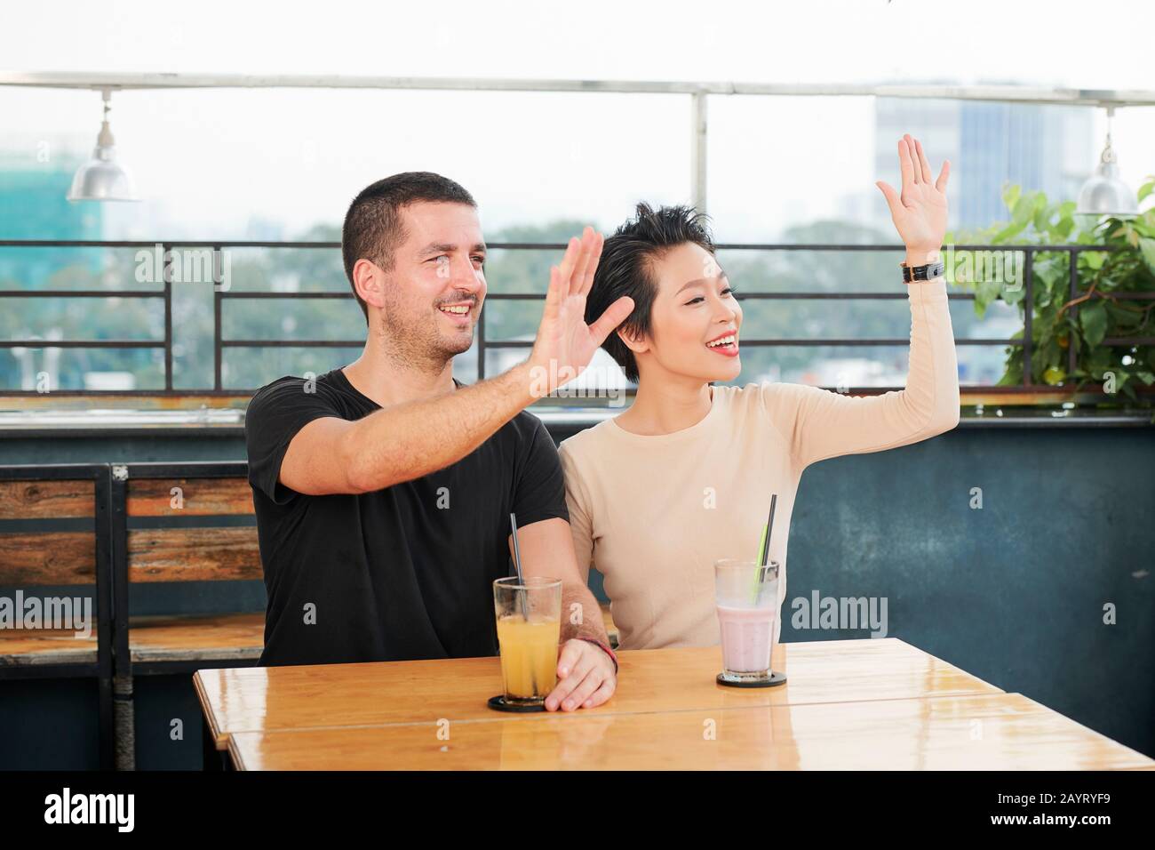 Smiling young people sitting at cafe table with cocktails and waving ...