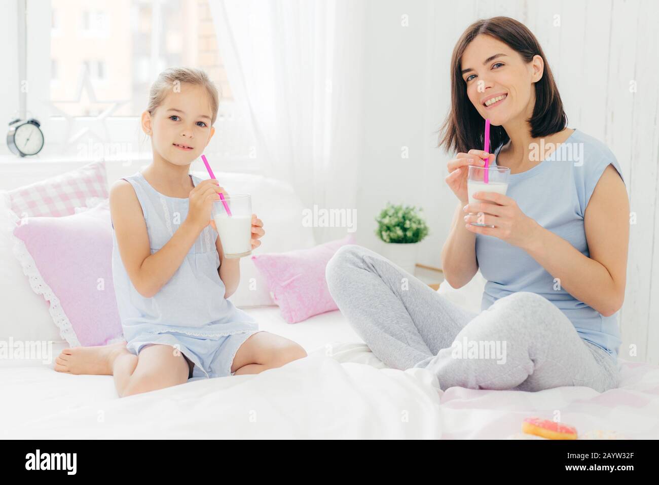 Happy brunette young mum and her pretty daughter sit on bed, drink milk ...