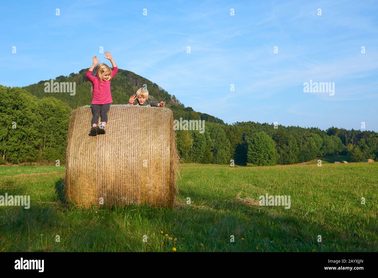 A young children boy and girl (siblings) playing on hay bale summertime ...