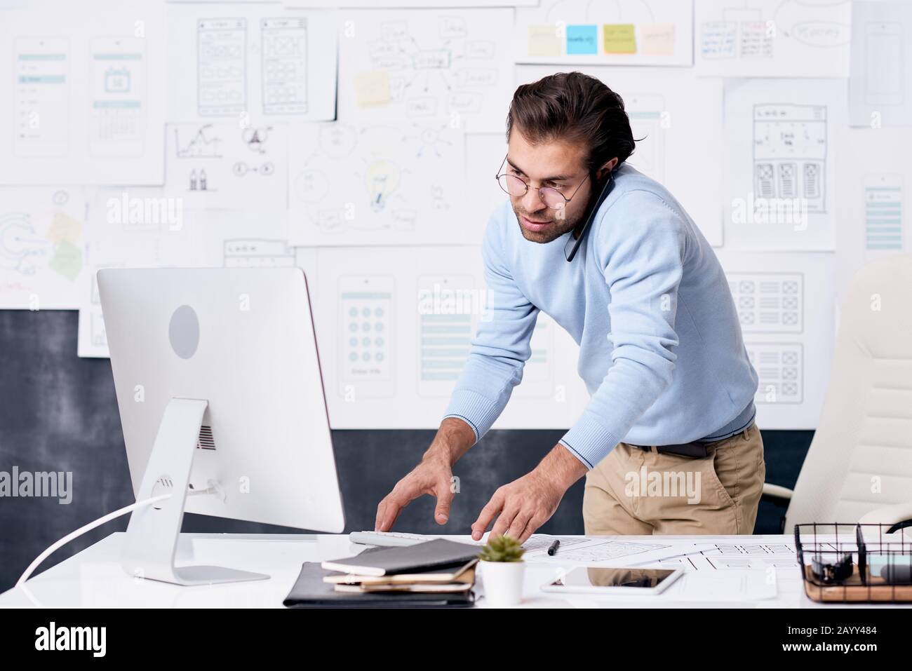 Horizontal portrait of busy young man standing at his desk working on ...