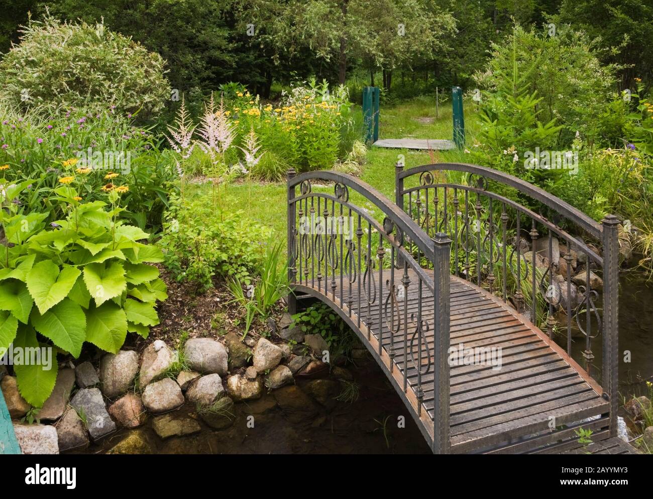 Wrought-iron footbridge over stream and rock edged border planted with ...