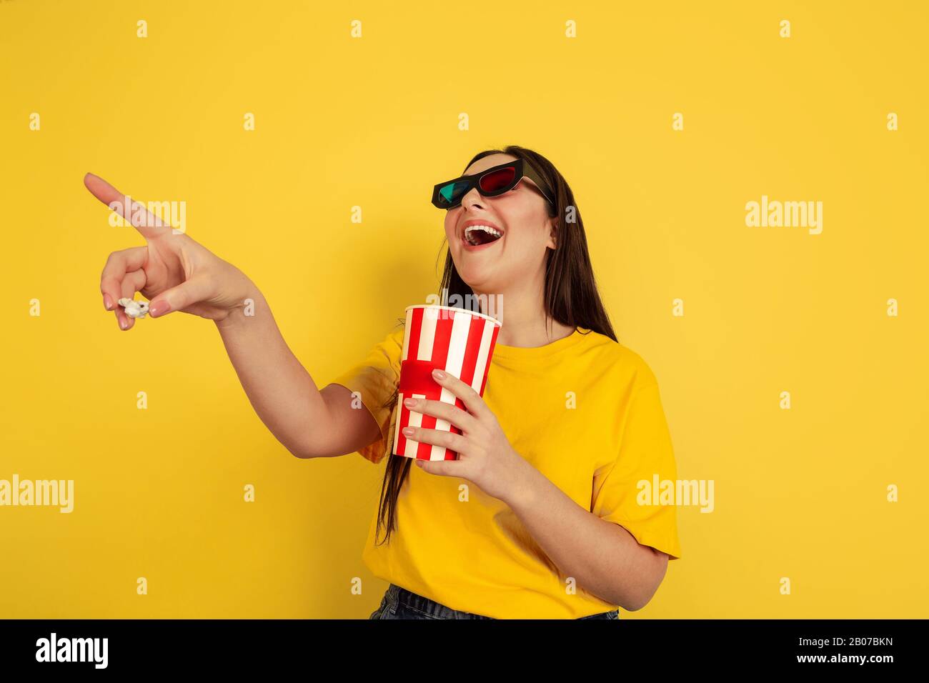 Watching cinema in 3D-eyewear with popcorn. Caucasian woman on yellow studio background ...