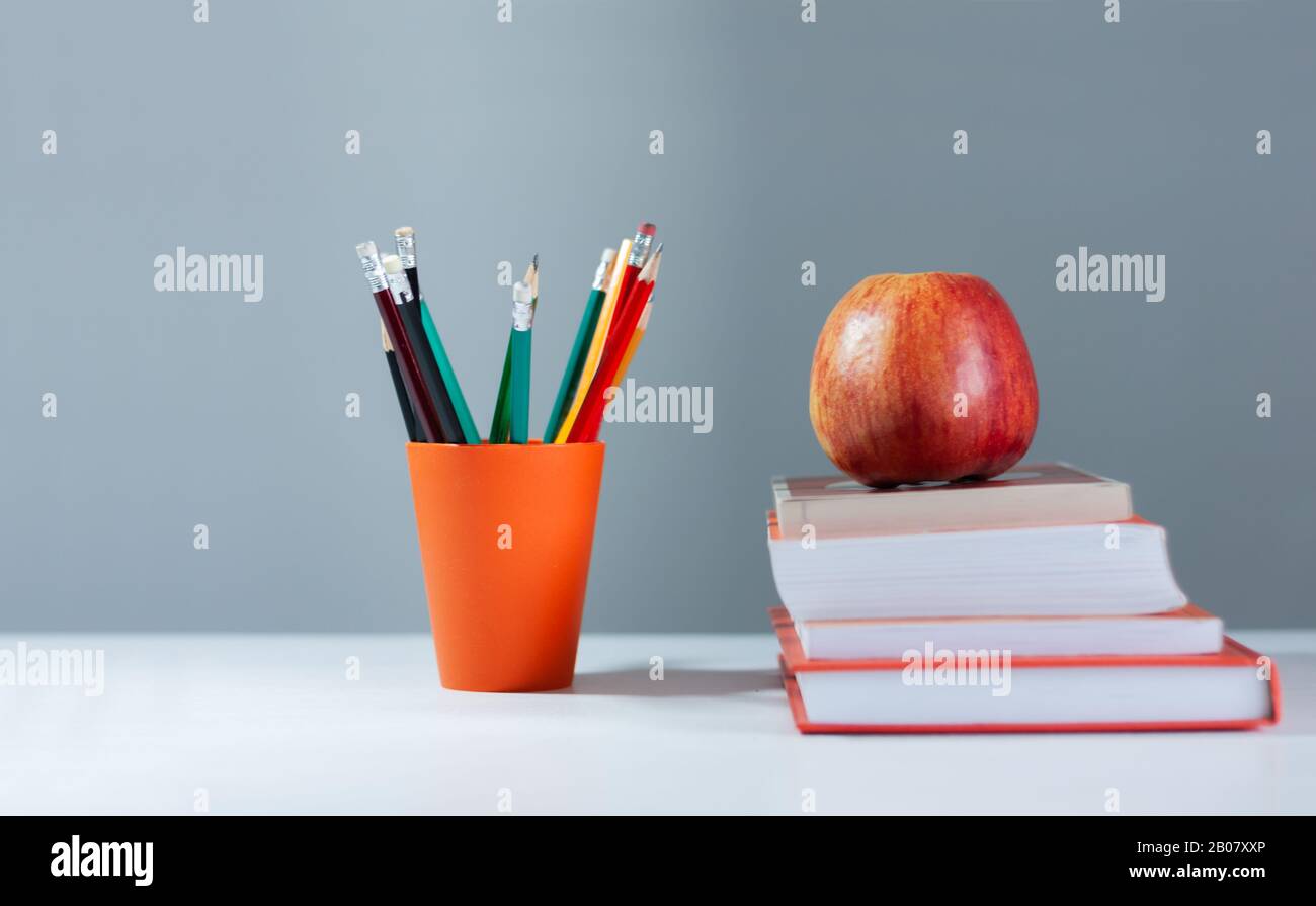 Orange pencil holder, stack of books on white table with red apple ...