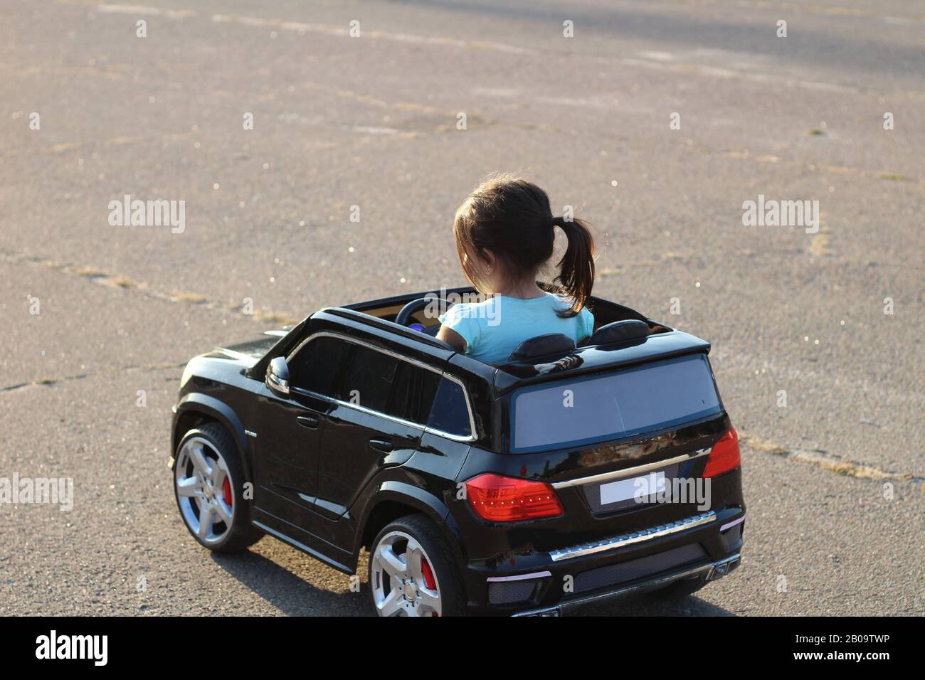 little girl with a tail drives a toy black car on asphalt in a square ...