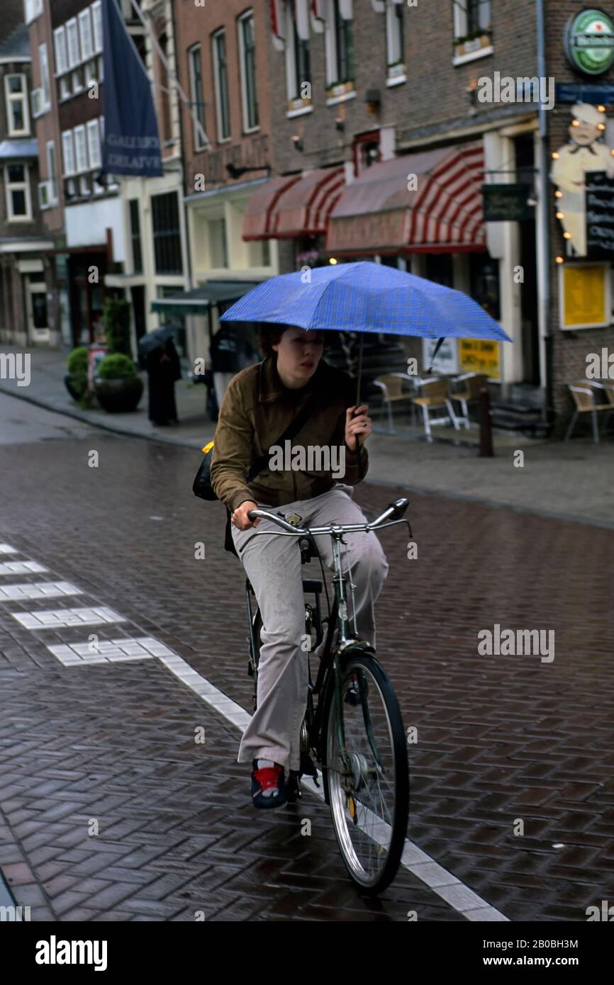 NETHERLANDS, HOLLAND, AMSTERDAM, WOMAN WITH UMBRELLA ON BICYCLE IN RAIN ...