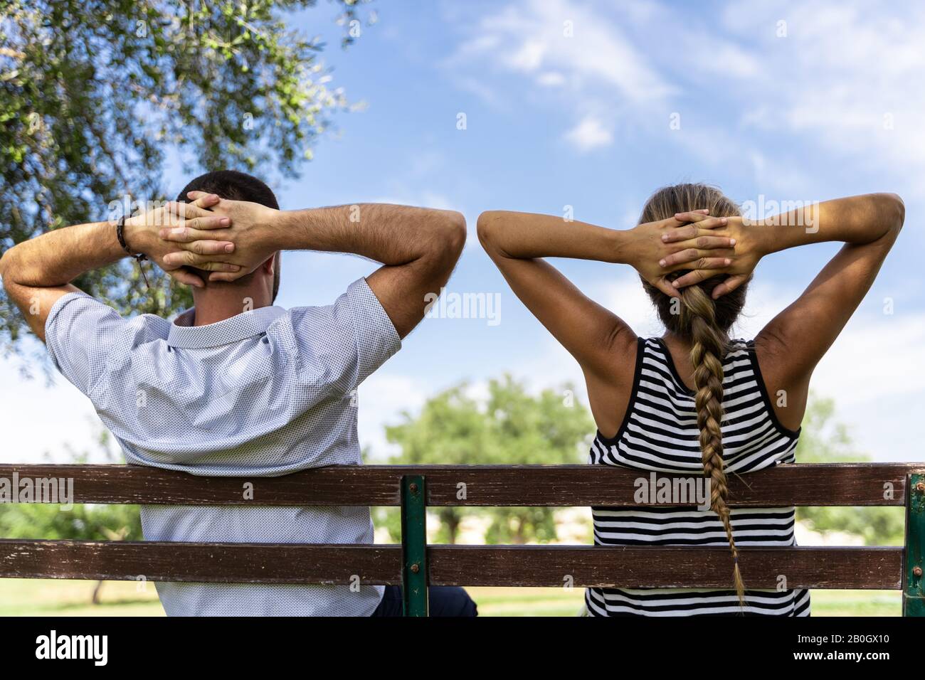 A young couple sitting on a wooden bench under a tree with their arms ...
