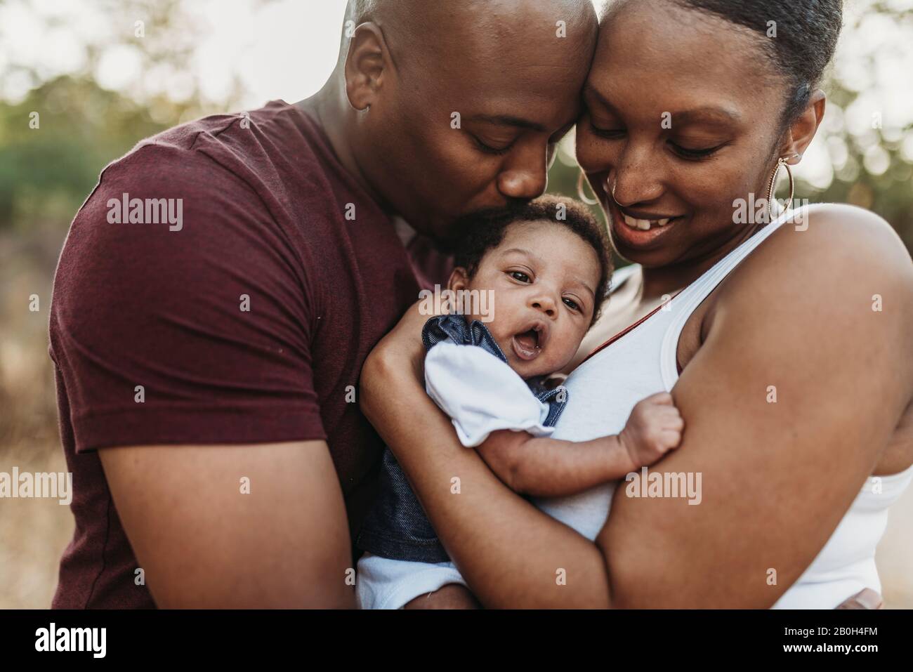 Close up happy mother and father cuddling infant girl backlit field ...