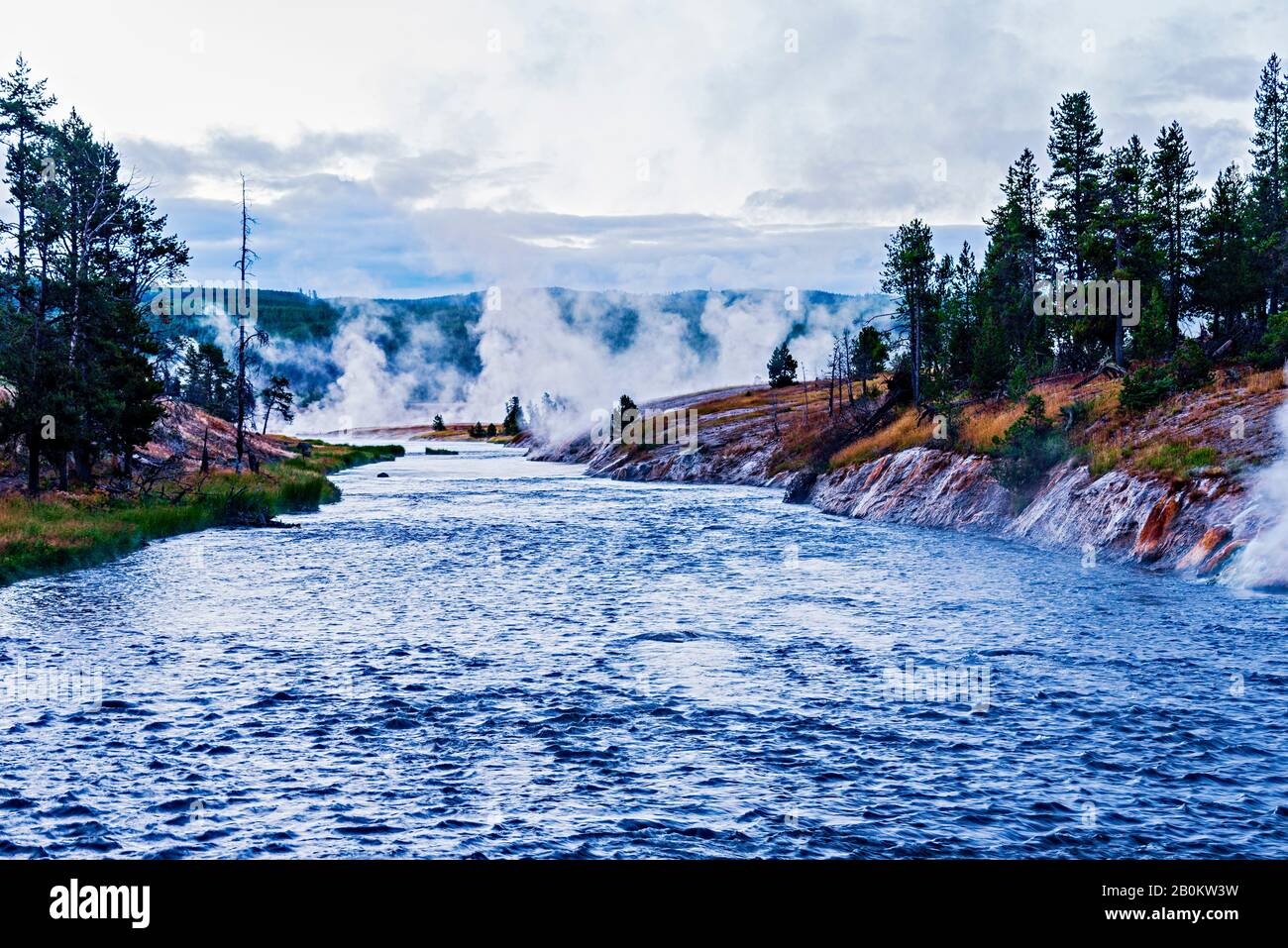 Blue river flowing from geysers and hills with green pine trees on both ...
