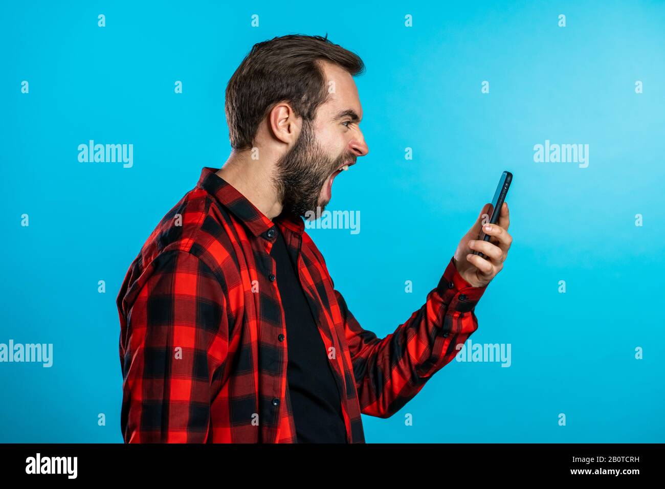 Angry modern hipster in red plaid shirt screaming down his mobile phone ...