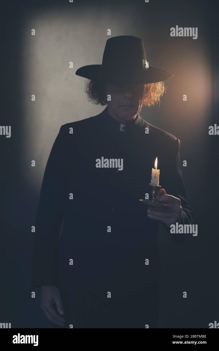 Mysterious victorian priest in black coat and hat holding candlestick ...