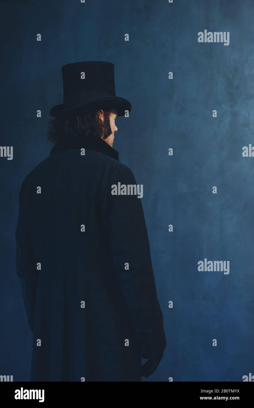 Edwardian man in long black coat and hat standing towards grey wall ...