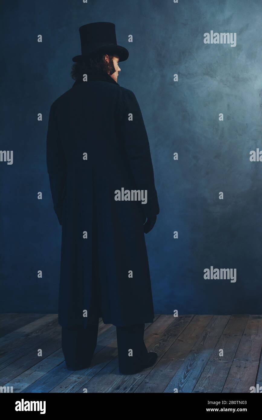 Edwardian man in long black coat and hat standing towards grey wall ...