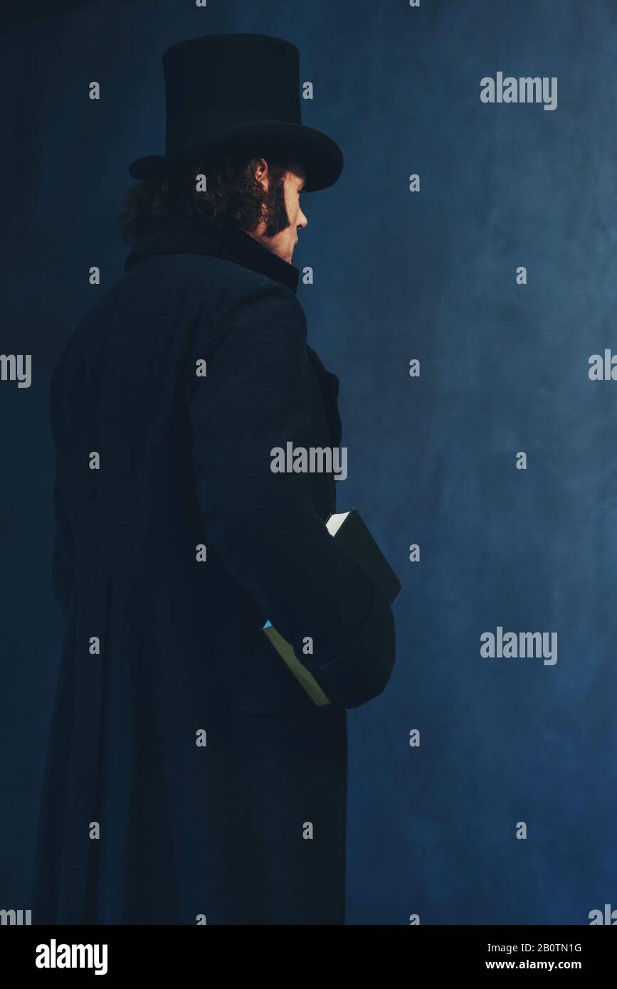 Edwardian man in long black coat and hat holding book standing towards ...