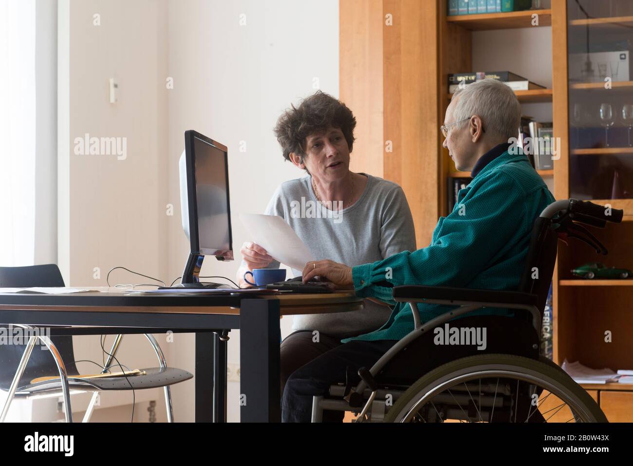 Senior woman sitting in a wheelchair at a computer, helper assisting ...