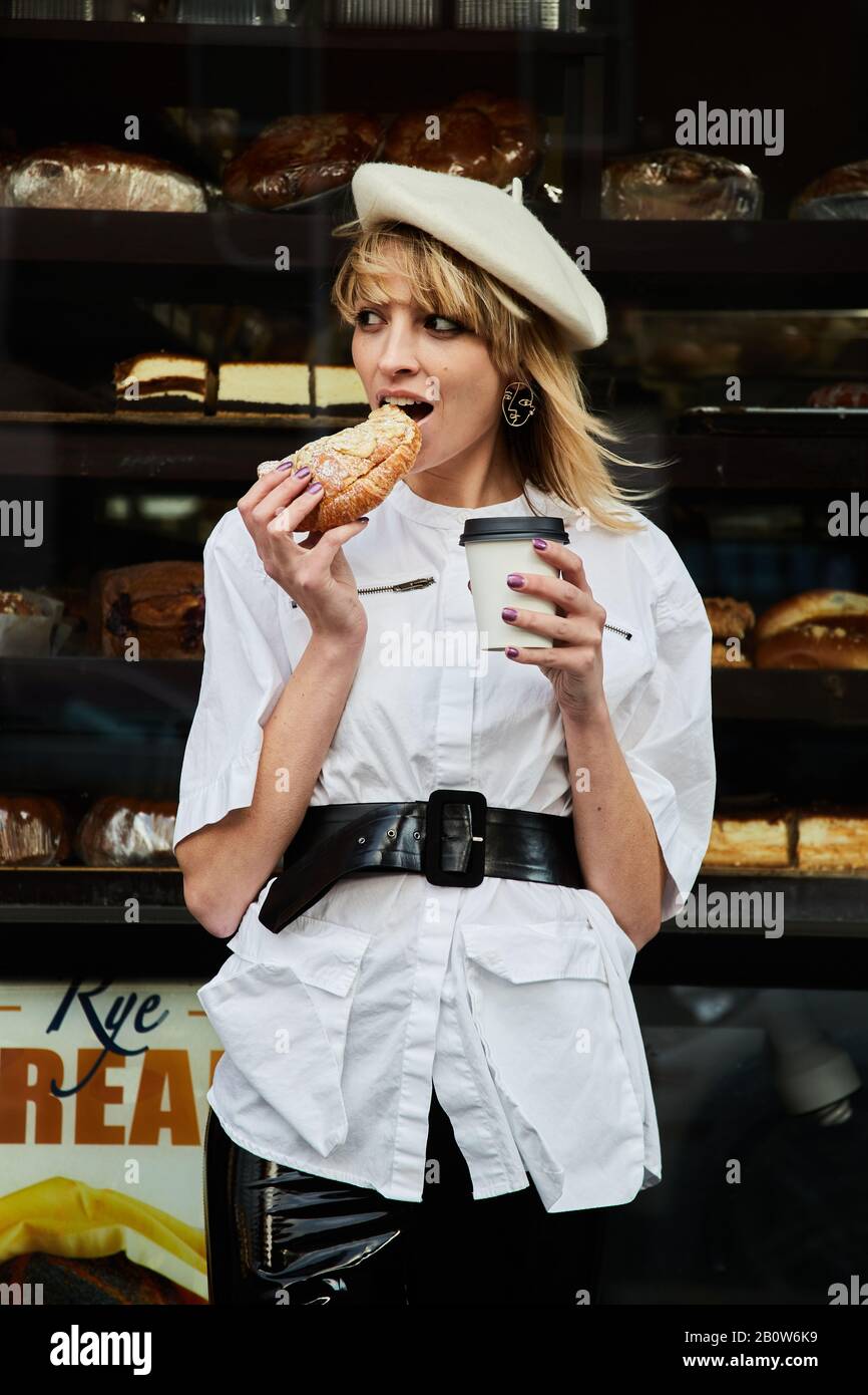 Pretty blonde woman having a pastry snack on the street, wearing a chic ...